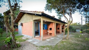 Uma casa térrea de tijolos com varanda coberta, chaminé visível e um pequeno jardim com árvores e arbustos.