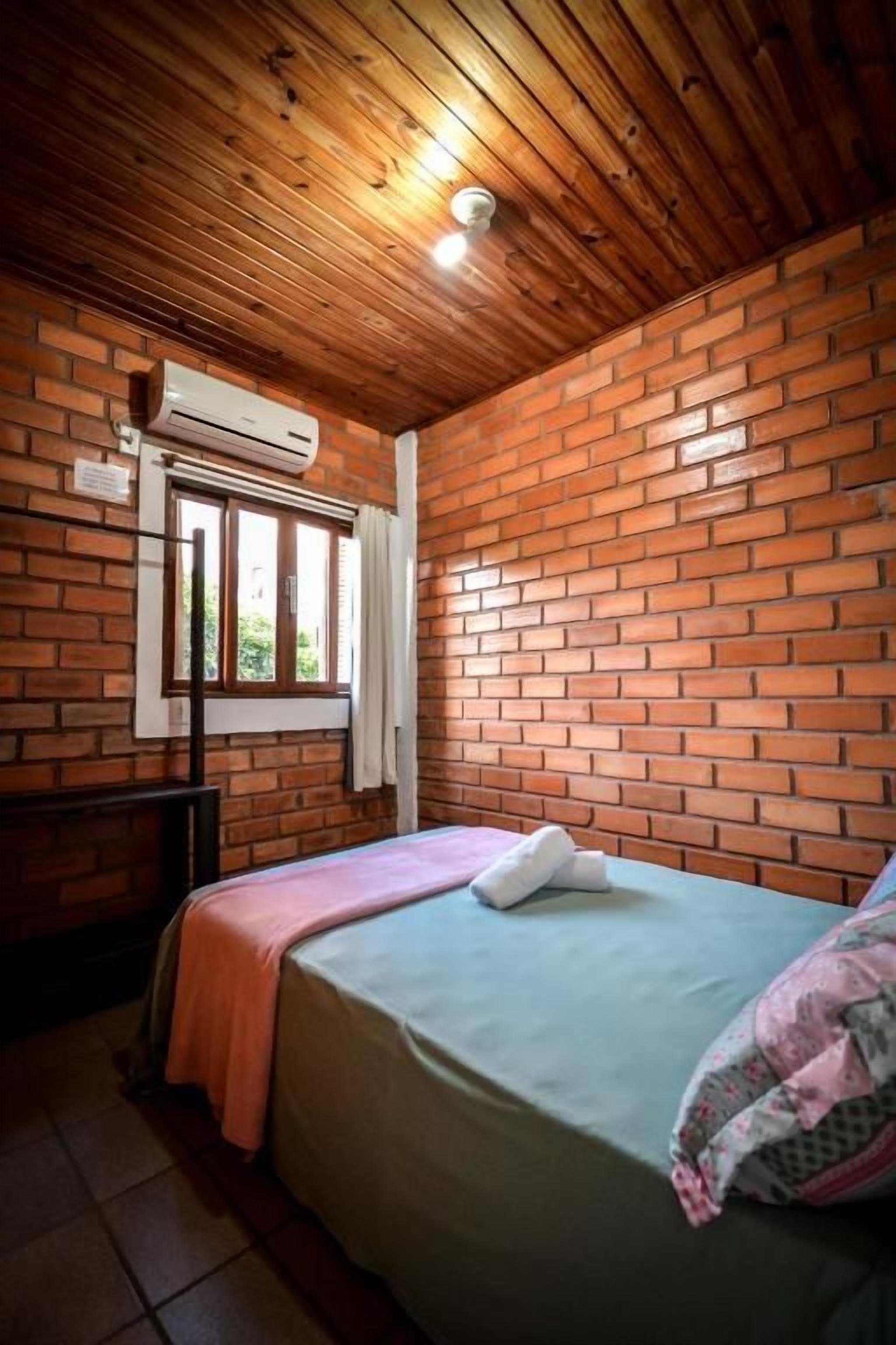 Um quarto com parede de tijolos, cama com colcha verde, janela com cortina branca e teto com vigas de madeira.