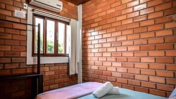 Um quarto com parede de tijolos, cama com colcha verde, janela com cortina branca e teto com vigas de madeira.