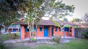 Uma casa de tijolos de dois andares com telhado vermelho, porta azul e um pequeno jardim frontal com uma árvore e um banco.