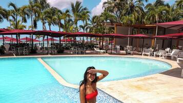 Uma mulher de biquíni vermelho em uma piscina azul cristalina, com palmeiras e guarda-sóis vermelhos ao fundo.