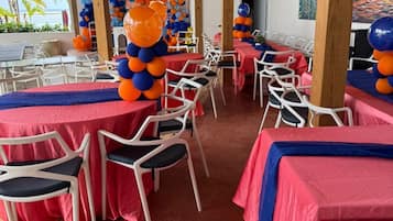 Uma área de refeições ao ar livre com mesas cobertas por toalhas vermelhas, cadeiras brancas e um deck de madeira.