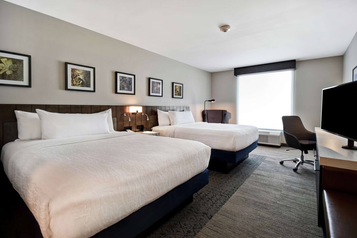 Quarto de hotel com duas camas, uma escrivaninha com televisão, uma cadeira e quadros nas paredes.