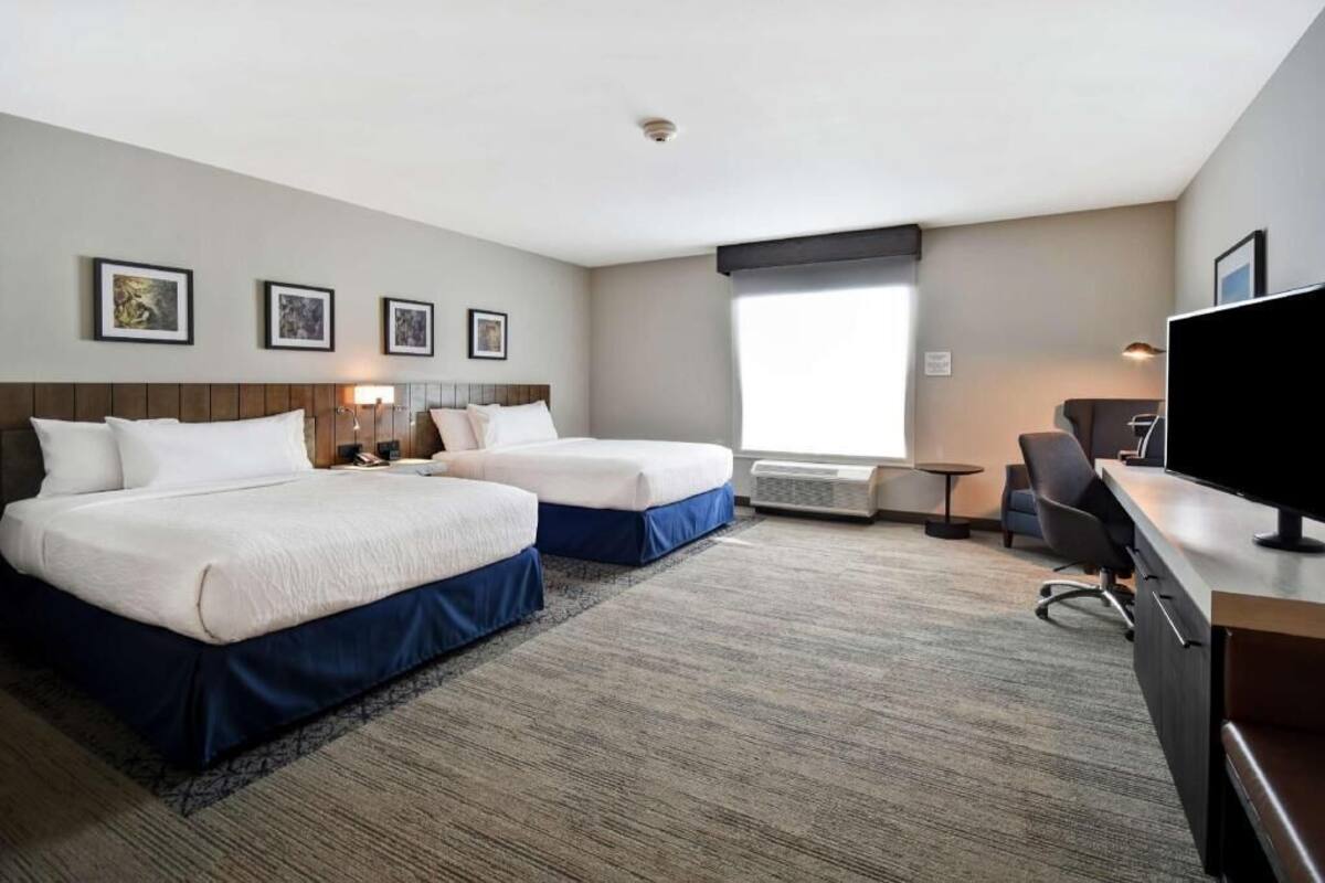 Quarto de hotel com duas camas, uma escrivaninha, uma cadeira, uma TV e quadros nas paredes.