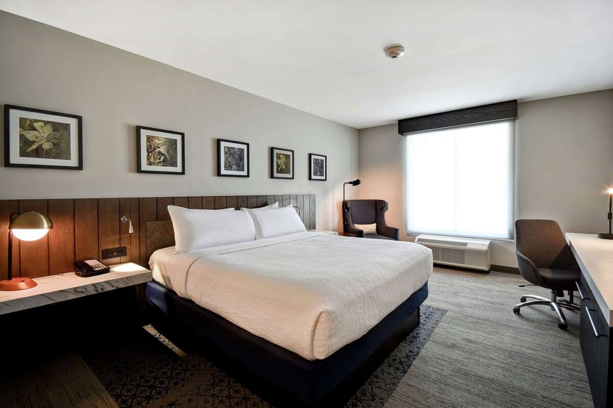 Quarto de hotel com cama, escrivaninha, cadeira e quadros na parede.