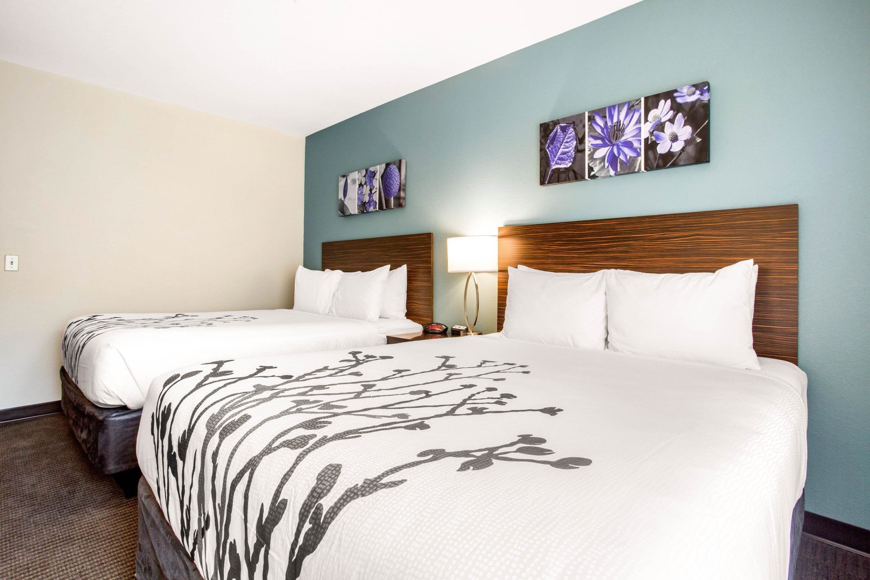 Quarto de hotel com duas camas, cabeceira de madeira, mesa de cabeceira com abajur e uma obra de arte floral na parede.