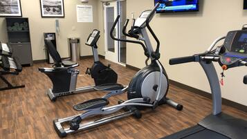 Centro de fitness com máquinas de cardio, televisão e relógio na parede.