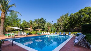 Piscina retangular com deck ladrilhado, rodeada por espreguiçadeiras e guarda-sóis, inserida em um jardim paisagístico com palmeiras e outras plantas.