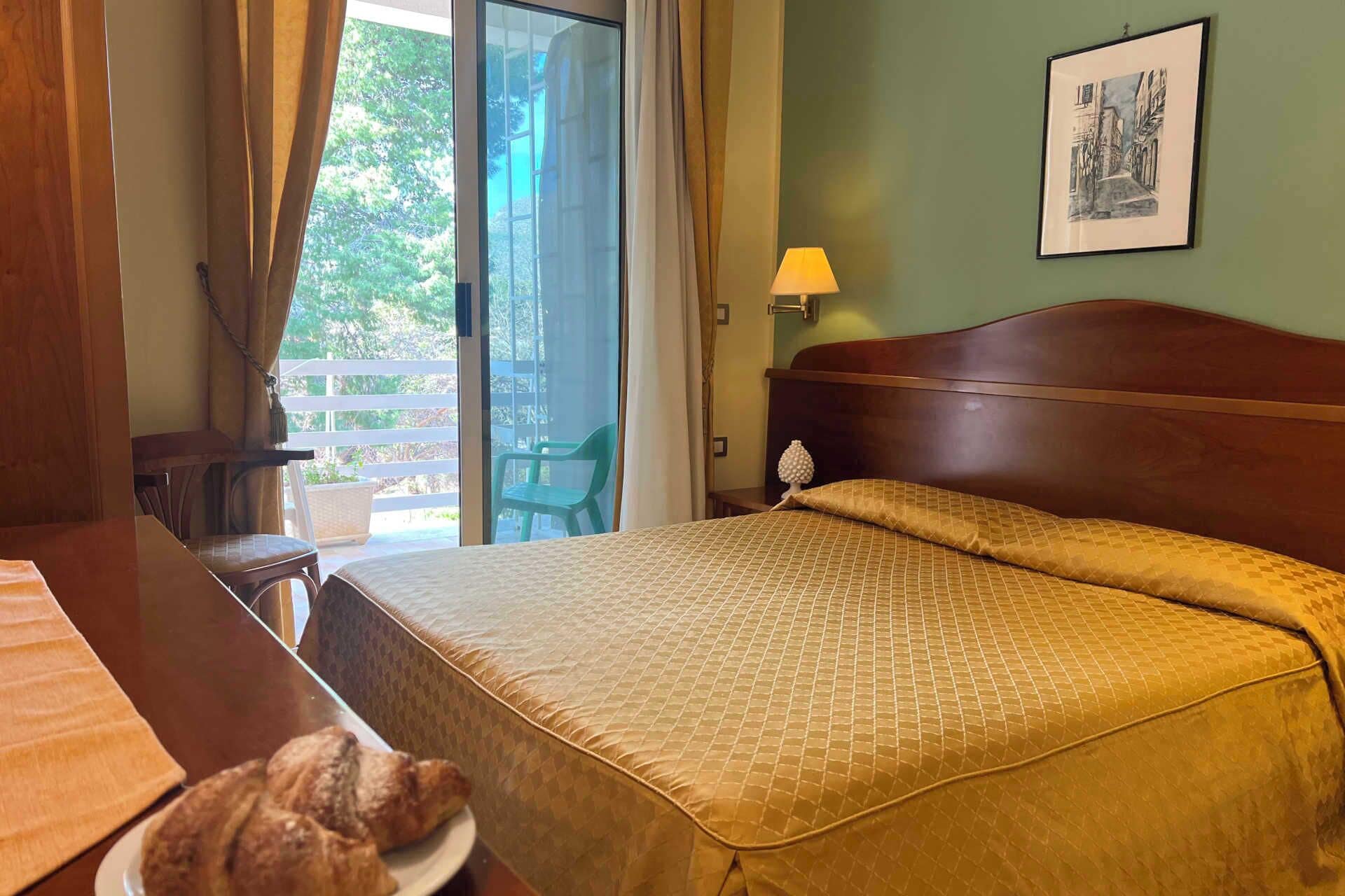 Quarto de hotel com cama, escrivaninha, cadeira, luminária e vista para o exterior.