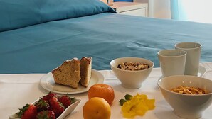 Cama arrumada com cabeceira azul, mesa de cabeceira com abajur, e bandeja de café da manhã com pão, morangos, laranja e tigelas de cereal e iogurte.
