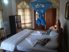 Quarto de hotel com cama, mesa de cabeceira, abajur, ventilador e frigorífico.