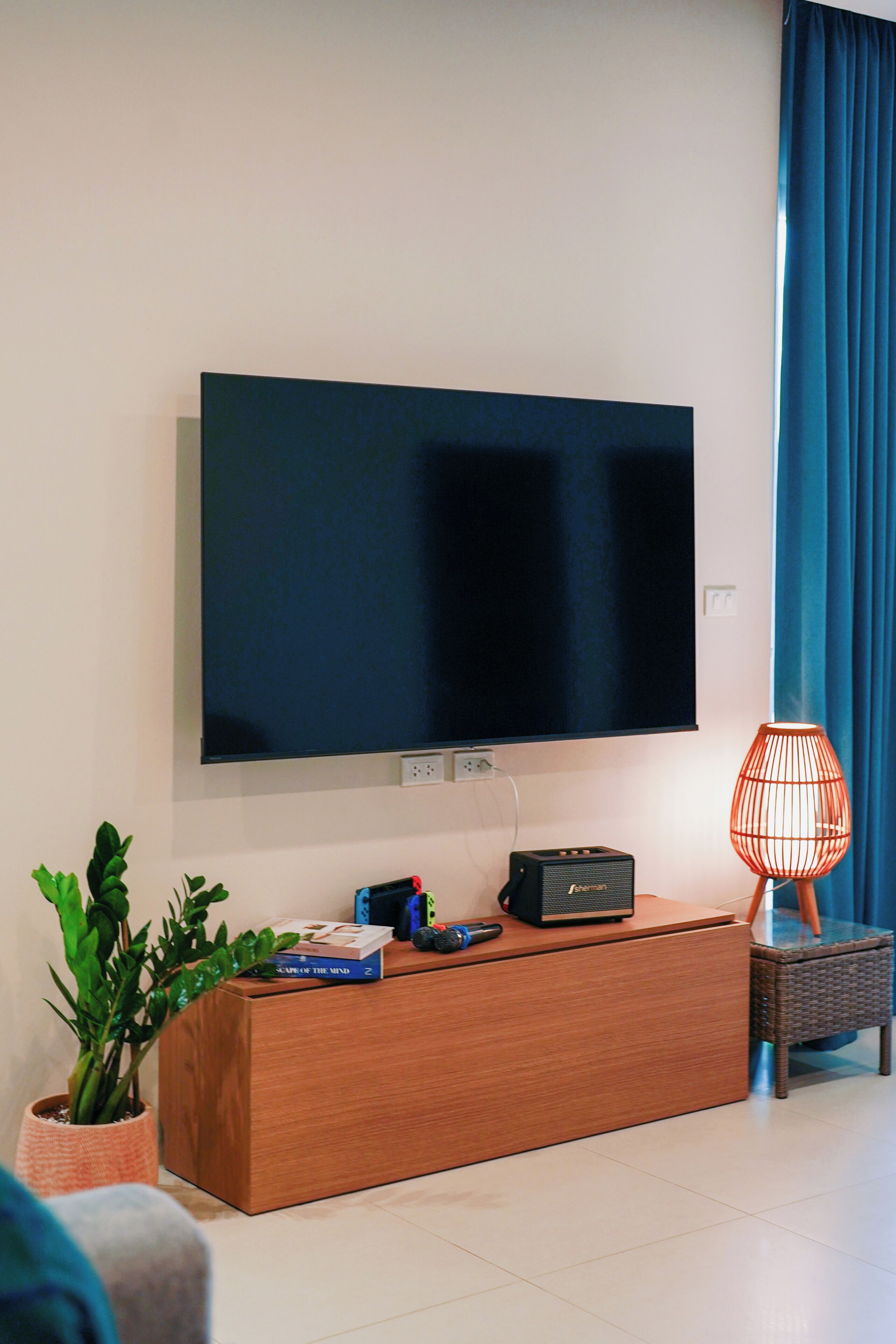 Uma sala de estar moderna com uma televisão de tela plana fixada na parede, um móvel para eletrônicos de madeira, um vaso de planta e uma mesinha de vime com um abajur.