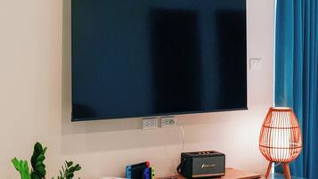 Uma sala de estar moderna com uma televisão de tela plana fixada na parede, um móvel para eletrônicos de madeira, um vaso de planta e uma mesinha de vime com um abajur.