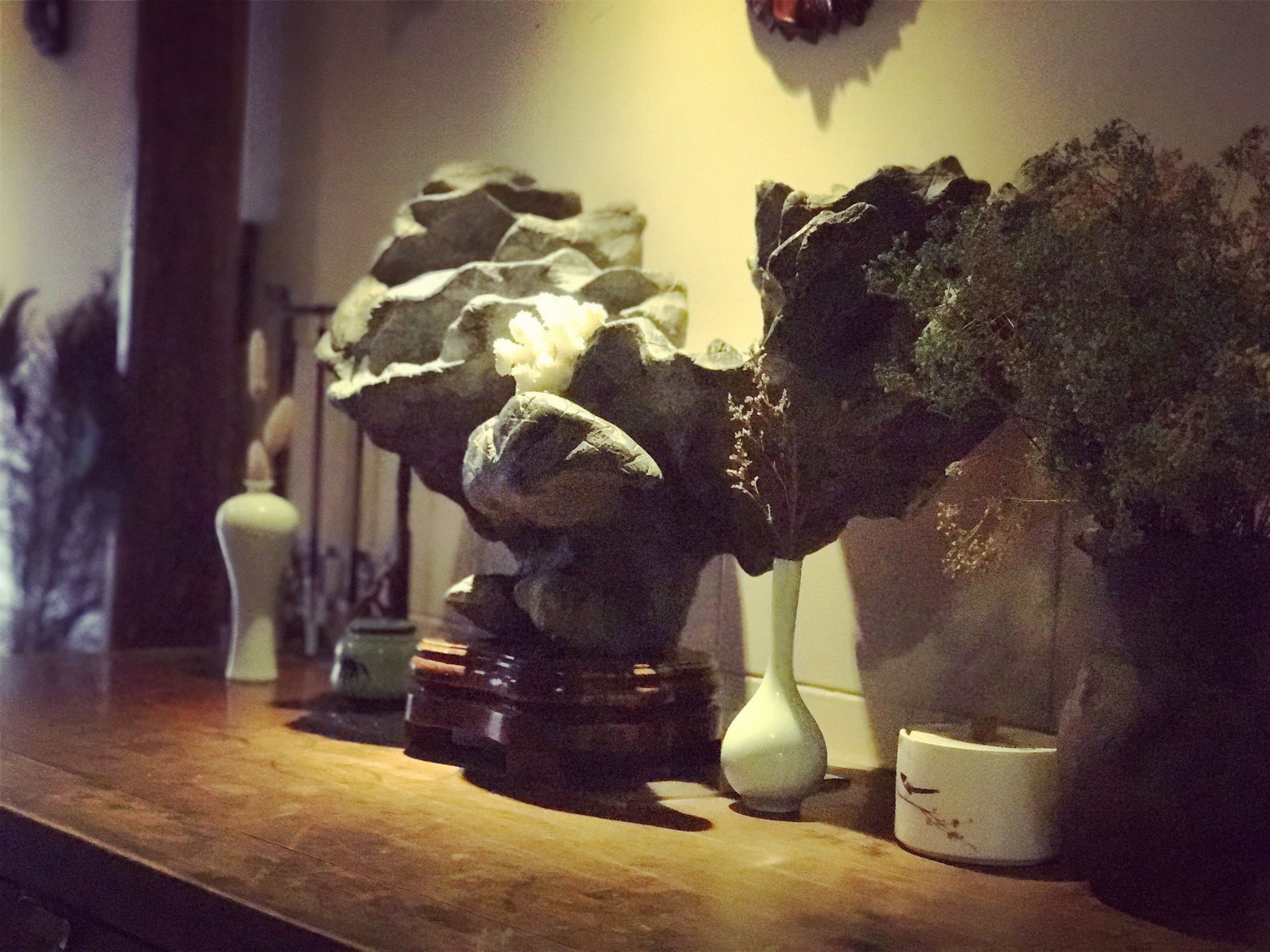 Uma prateleira de madeira com diversos objetos decorativos, incluindo uma escultura de pedra, um vaso com planta e uma pequena xícara branca.