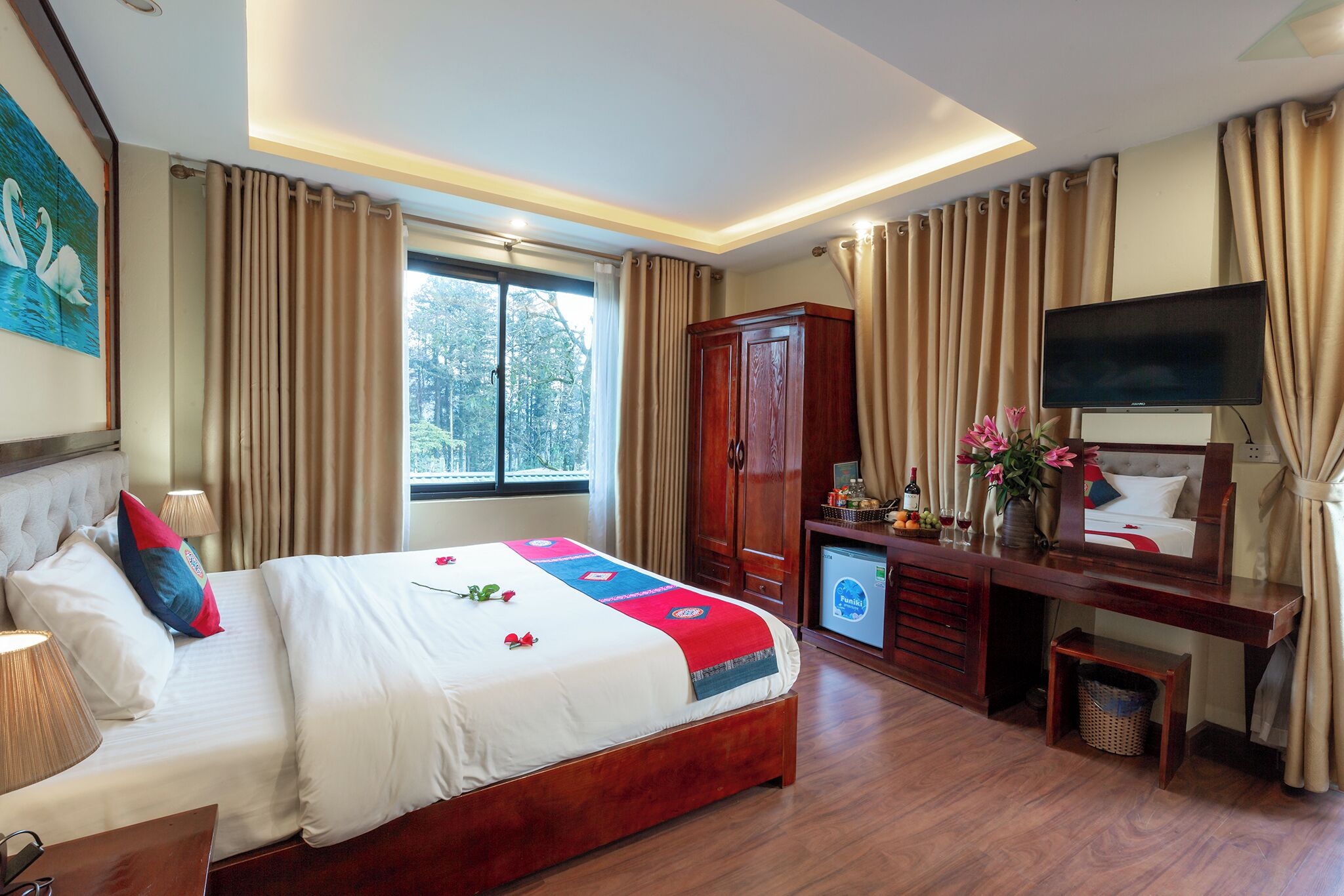 Quarto de hotel com uma cama grande, uma escrivaninha com televisão, um minibar e vista para árvores pela janela.