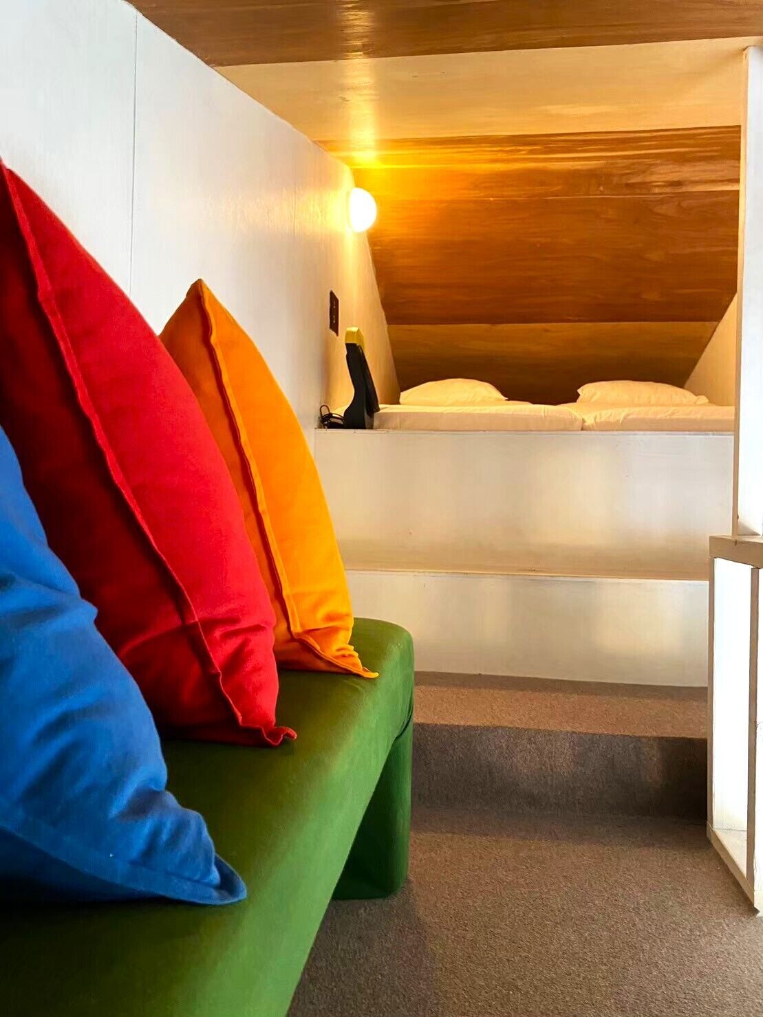 Quarto pequeno e acolhedor, com uma cama beliche, um sofá verde e almofadas coloridas, além de teto de madeira.