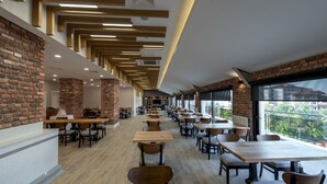 Um restaurante amplo com mesas e cadeiras compridas, paredes de tijolo e grandes janelas com vista para a cidade.