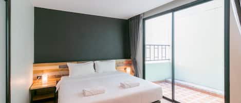 Quarto de hotel com uma cama grande, mesa de cabeceira com abajur, porta de vidro deslizante para uma varanda e uma parede escura.