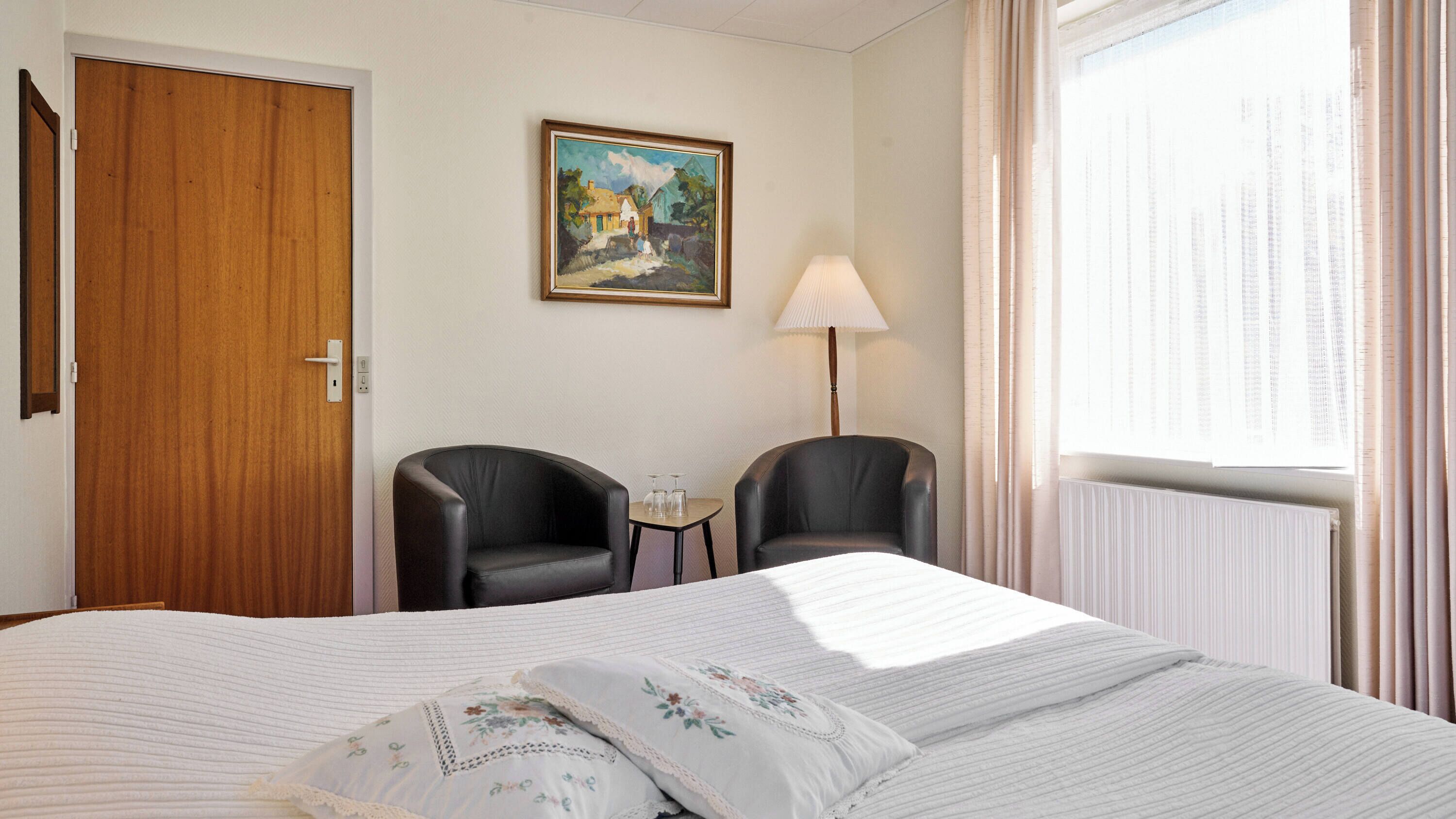 Quarto de hotel com cama, duas cadeiras, uma mesa com copos, um quadro, um abajur e uma janela com cortinas.