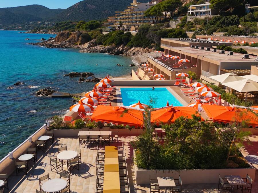 Um resort com piscina rodeada por espreguiçadeiras e guarda-sóis, com vista para uma área costeira com águas azuis cristalinas e colinas ao fundo.
