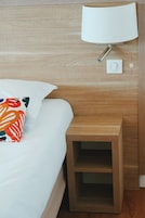 Cama bem arrumada com lençóis brancos e almofada colorida, mesa de cabeceira com abajur e luminária na parede.