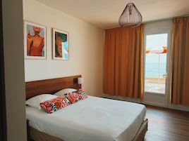 Quarto de hotel com cama, duas imagens emolduradas na parede, luminária pendente e vista para a praia pela janela.