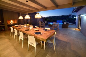 Uma área de refeições ao ar livre com uma mesa de madeira posta para seis pessoas, cadeiras brancas e luminárias pendentes.