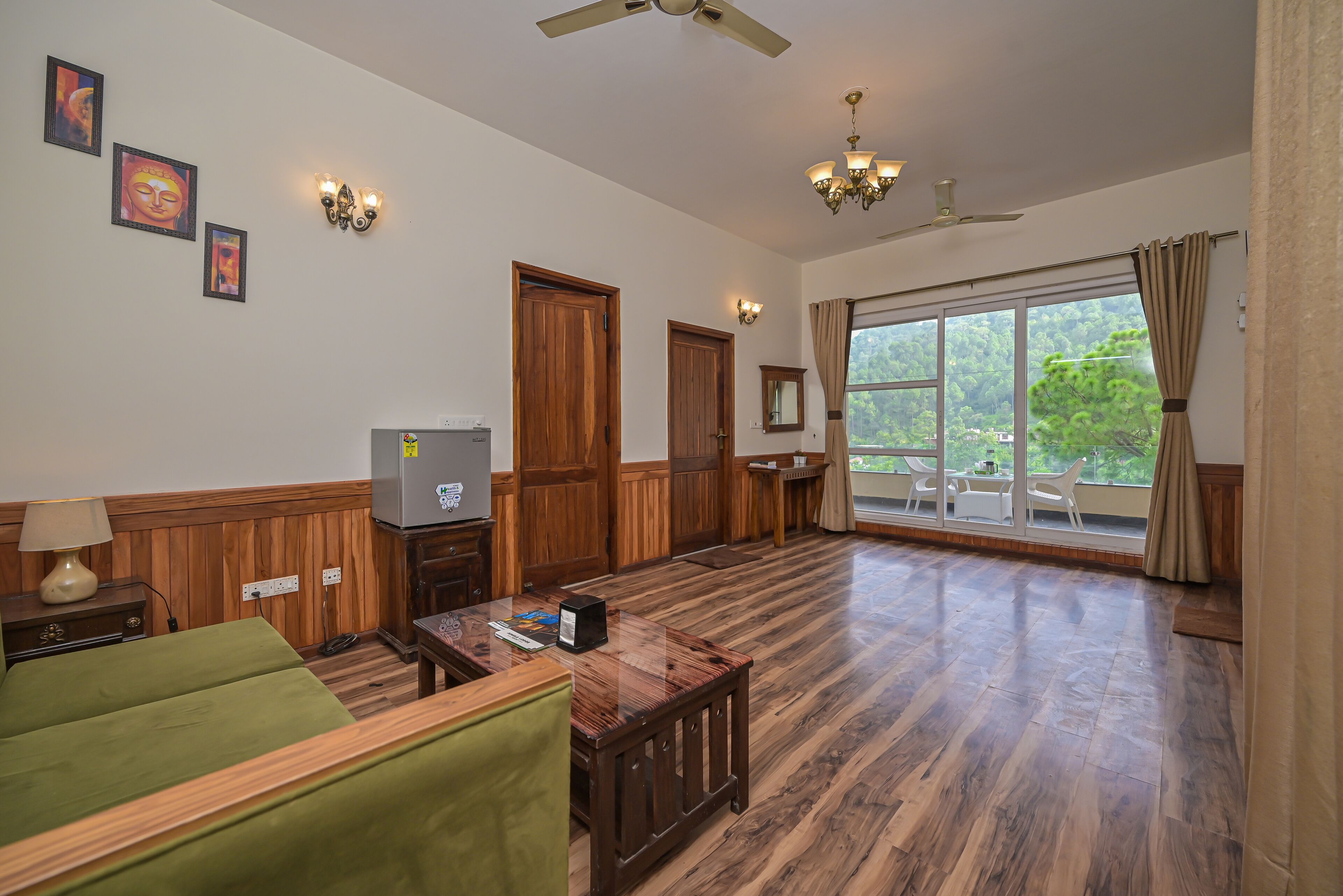 Uma sala de estar ampla com piso de madeira, um sofá verde, uma mesa de centro de madeira e uma televisão. Há portas de madeira, uma janela com vista para o verde e um ventilador de teto.