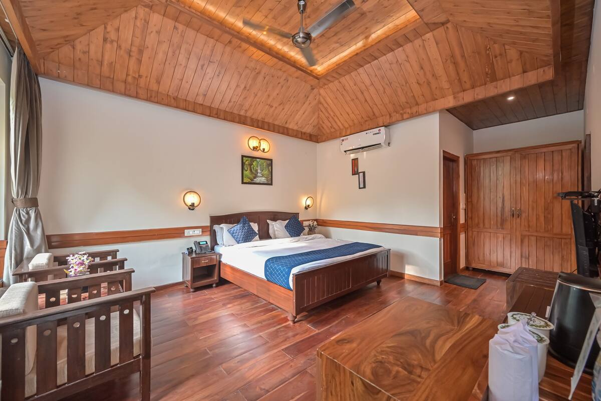 Um quarto espaçoso com teto de madeira, uma cama grande com colcha azul, duas mesas de cabeceira, um criado-mudo e um guarda-roupa de madeira.