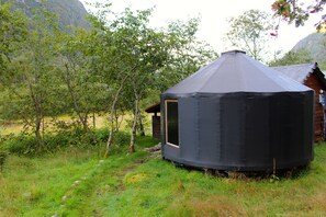 Uma yurt preta com porta de madeira, rodeada por grama e árvores, situada em uma paisagem natural.