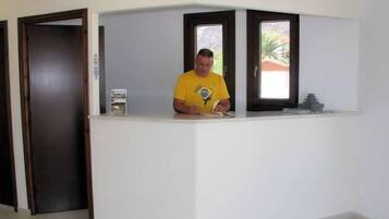 Um homem de camisa amarela escrevendo em uma mesa em um quarto com paredes brancas, uma janela e uma porta.