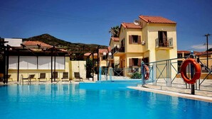 Piscina com espreguiçadeiras, um salva-vidas e um edifício com telhado de telhas vermelhas.