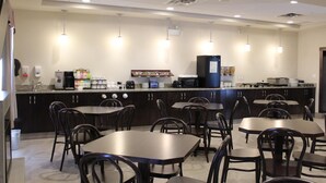 Cozinha de hotel moderna com um buffet, incluindo frigorífico, máquina de café e diversas opções de comida. Há várias mesas e cadeiras dispostas para refeições.