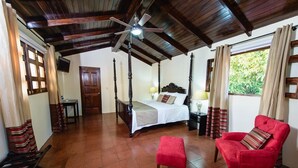 Quarto com cama de dossel, uma poltrona vermelha, um banquinho vermelho, um ventilador de teto e uma janela com cortinas.