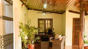 Uma área de estar acolhedora ao ar livre, com móveis de vime e uma mesa de vidro, rodeada por plantas em vasos e teto de madeira.