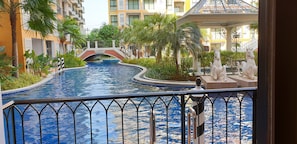 Piscina com uma ponte, rodeada por vegetação e palmeiras, com vista para edifícios altos ao fundo.