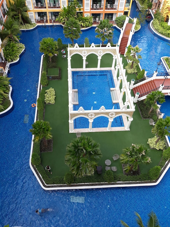 Uma piscina grande e central, rodeada por vegetação exuberante e palmeiras, com um edifício de vários níveis ao fundo.