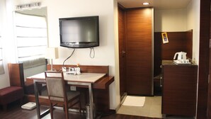 Quarto de hotel com uma escrivaninha de madeira, televisão, balança, cadeira, mesa pequena com um vaso, espelho e porta de madeira.