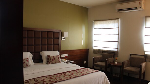 Quarto de hotel com uma cama grande, cabeceira de madeira, mesa de cabeceira com abajur, duas cadeiras, uma mesinha com um vaso e um aparelho de ar condicionado.