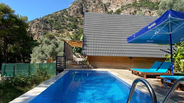 Uma piscina com deck revestido de azulejos, um edifício de madeira com telhado em azulejos e uma montanha ao fundo.