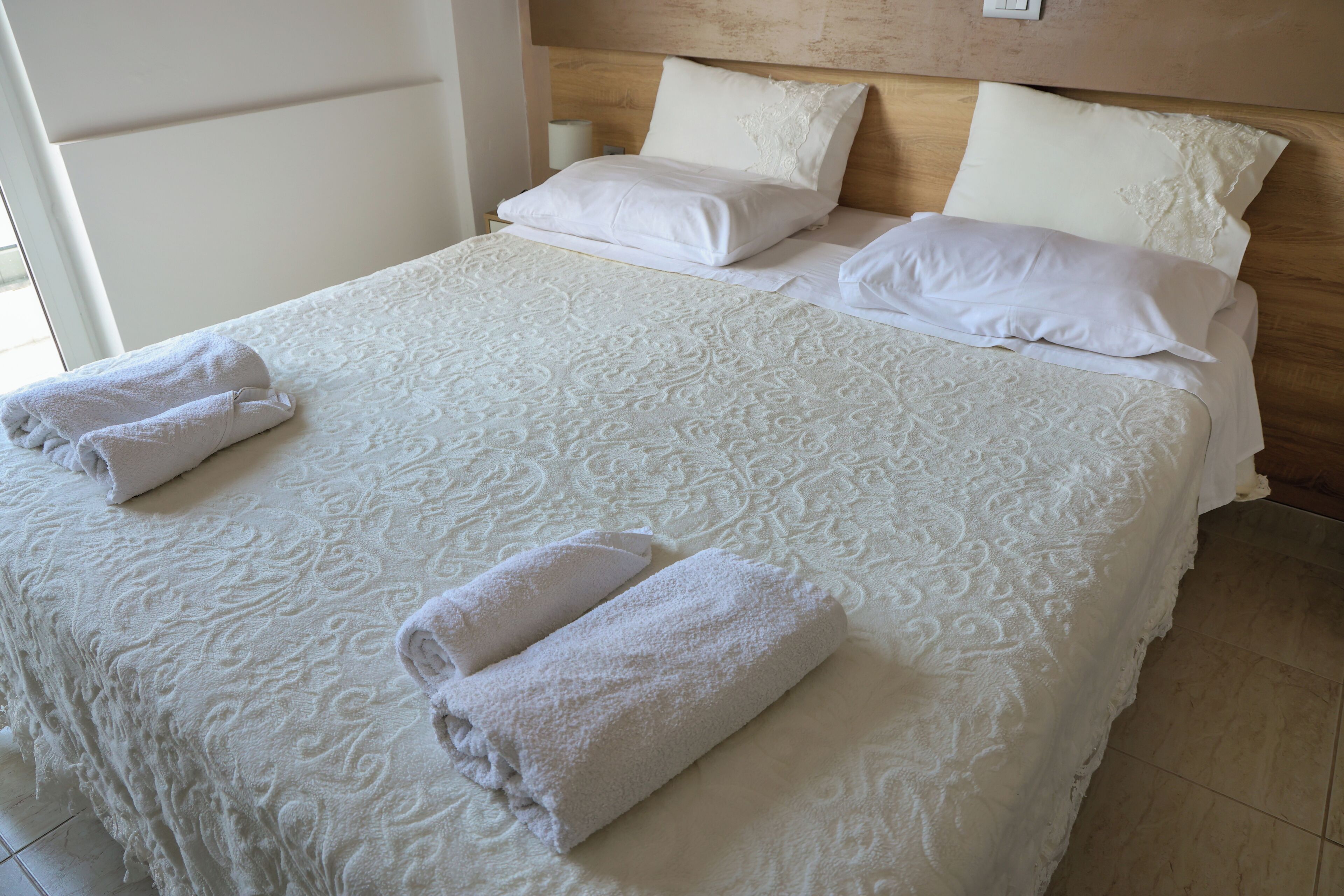 Cama bem arrumada com lençóis e toalhas brancos, cabeceira de madeira e parede lisa.
