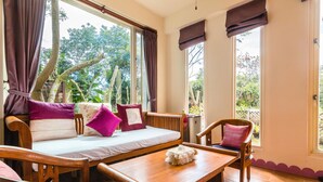 Um quarto acolhedor com sofá e mesa de madeira, e duas cadeiras. Há grandes janelas com cortinas, e vista para árvores lá fora.