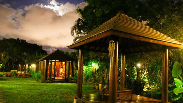 Um gazebo de madeira com telhado inclinado, rodeado por vegetação exuberante e um jardim bem iluminado à noite.