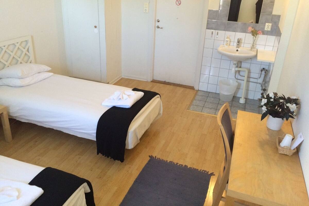 Quarto de hotel com duas camas, piso de madeira, uma mesa pequena com uma cadeira, pia e espelho.