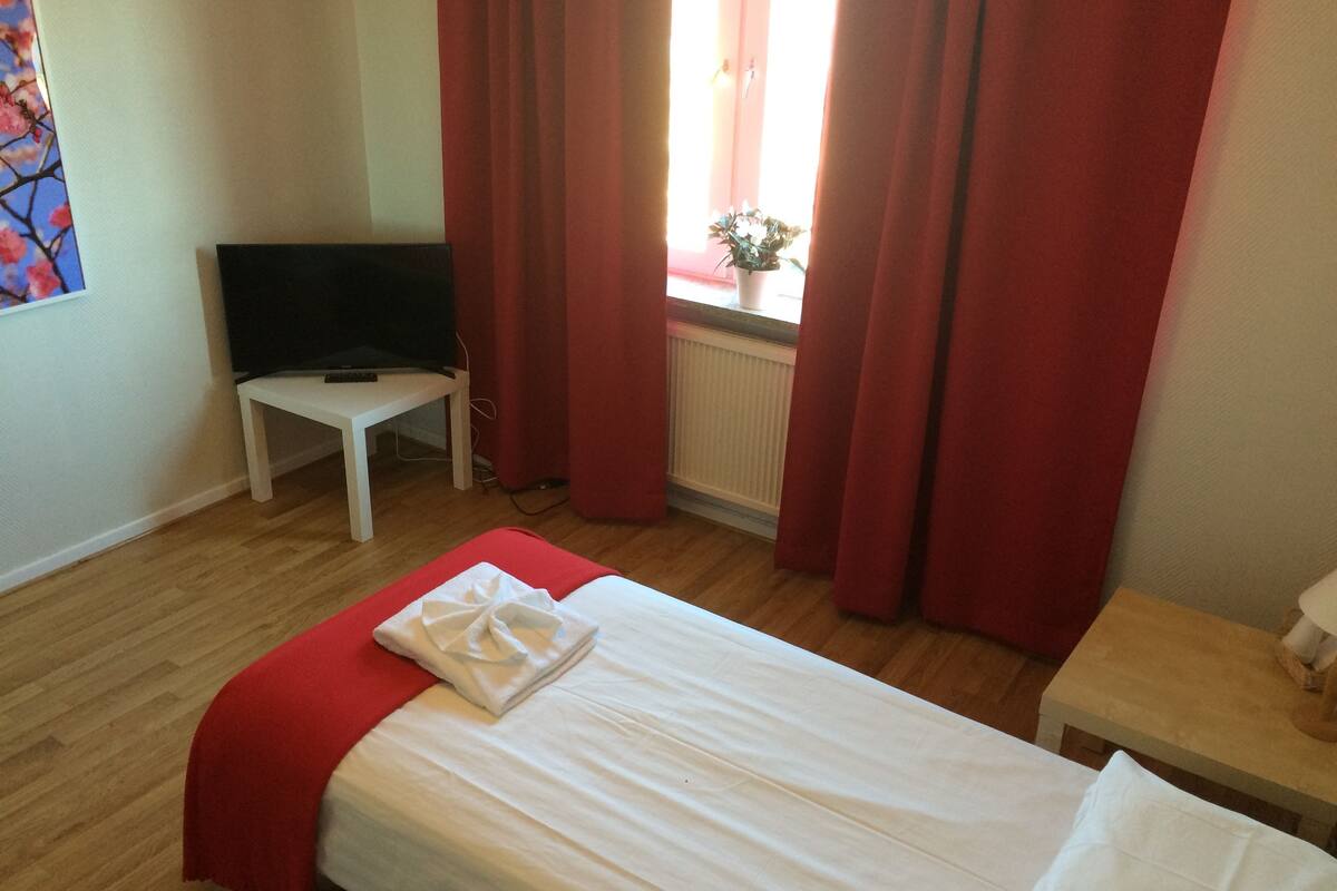 Quarto de hotel com cama, uma mesa pequena, televisão e cortinas vermelhas.