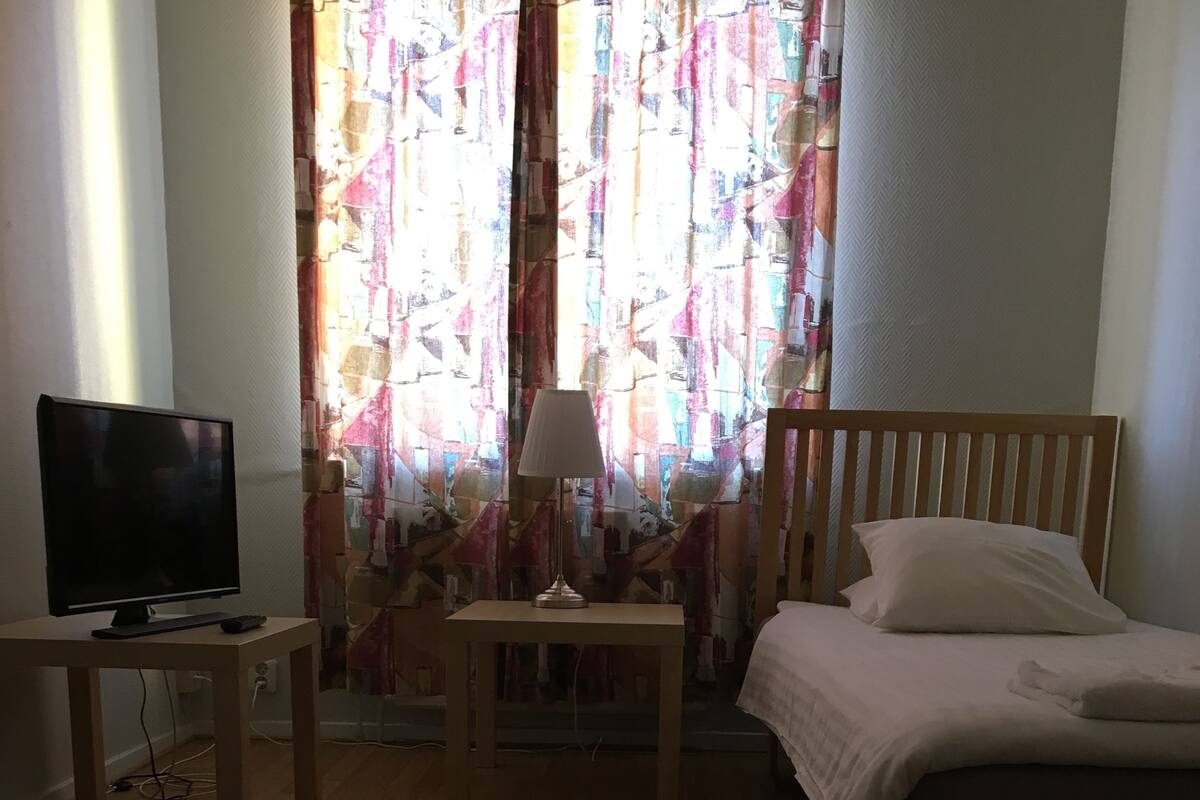 Quarto com cama, mesa de cabeceira com abajur, televisão em móvel e janela com cortinas.