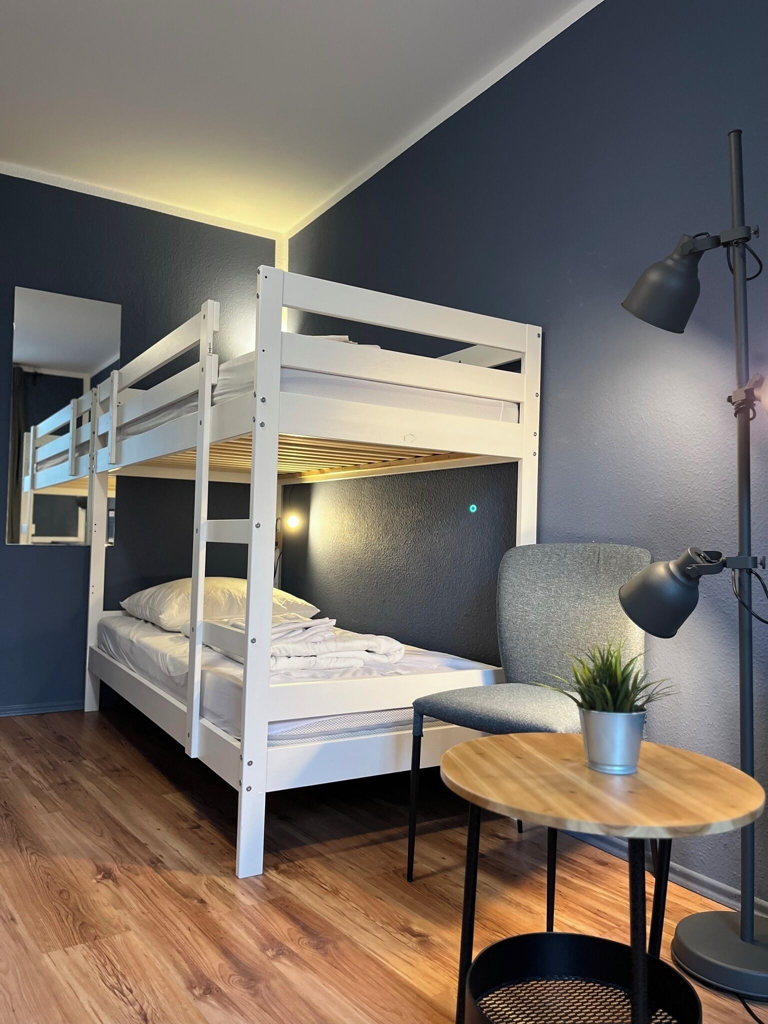 Uma cama beliche com escrivaninha e cadeira, uma mesinha pequena, um abajur e um vaso de planta num quarto com piso de madeira e paredes escuras.