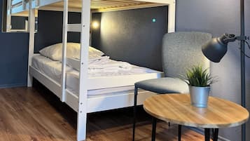 Uma cama beliche com escrivaninha e cadeira, uma mesinha pequena, um abajur e um vaso de planta num quarto com piso de madeira e paredes escuras.