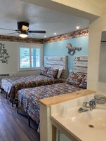 Quarto com duas camas, um ventilador de teto, uma janela com persianas e um cavalete decorativo na parede.