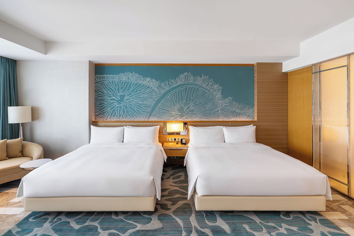 Quarto de hotel com duas camas, uma mesa de cabeceira com abajur, um sofá e uma grande obra de arte abstrata na parede.